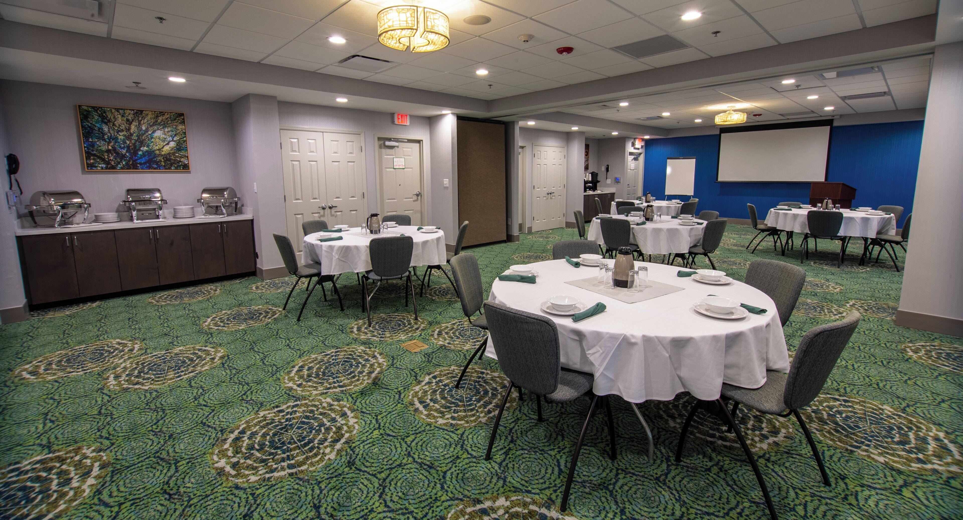 Sala de conferências com mesas redondas, cadeiras e área de buffet com equipamentos de serviço de alimentação.