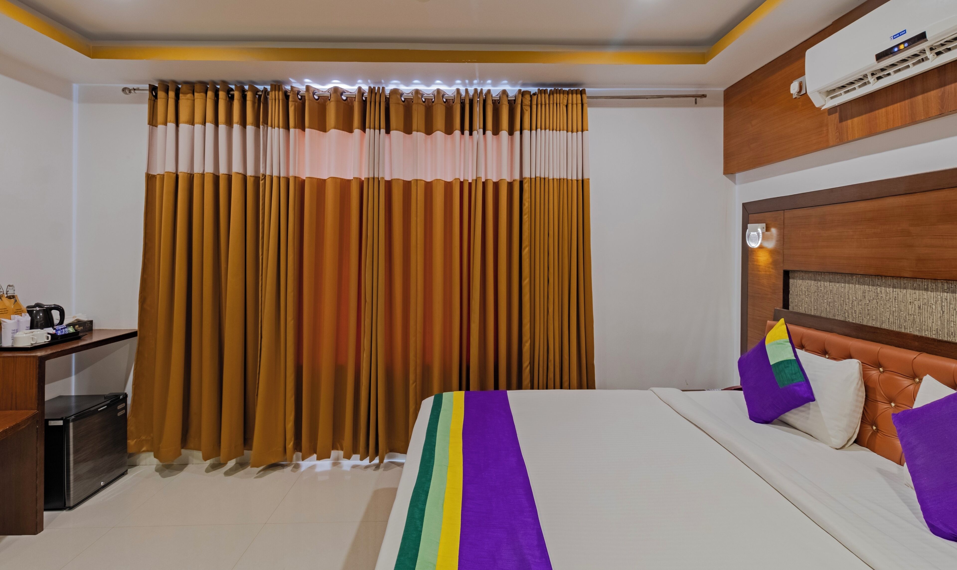 Quarto de hotel com cama, mesa de cabeceira, cafeteira e janela com cortinas.