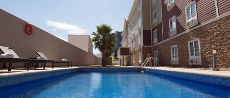 Piscina com espreguiçadeiras e uma palmeira, cercada por edifícios de vários andares com fachadas de tijolo e pedra.