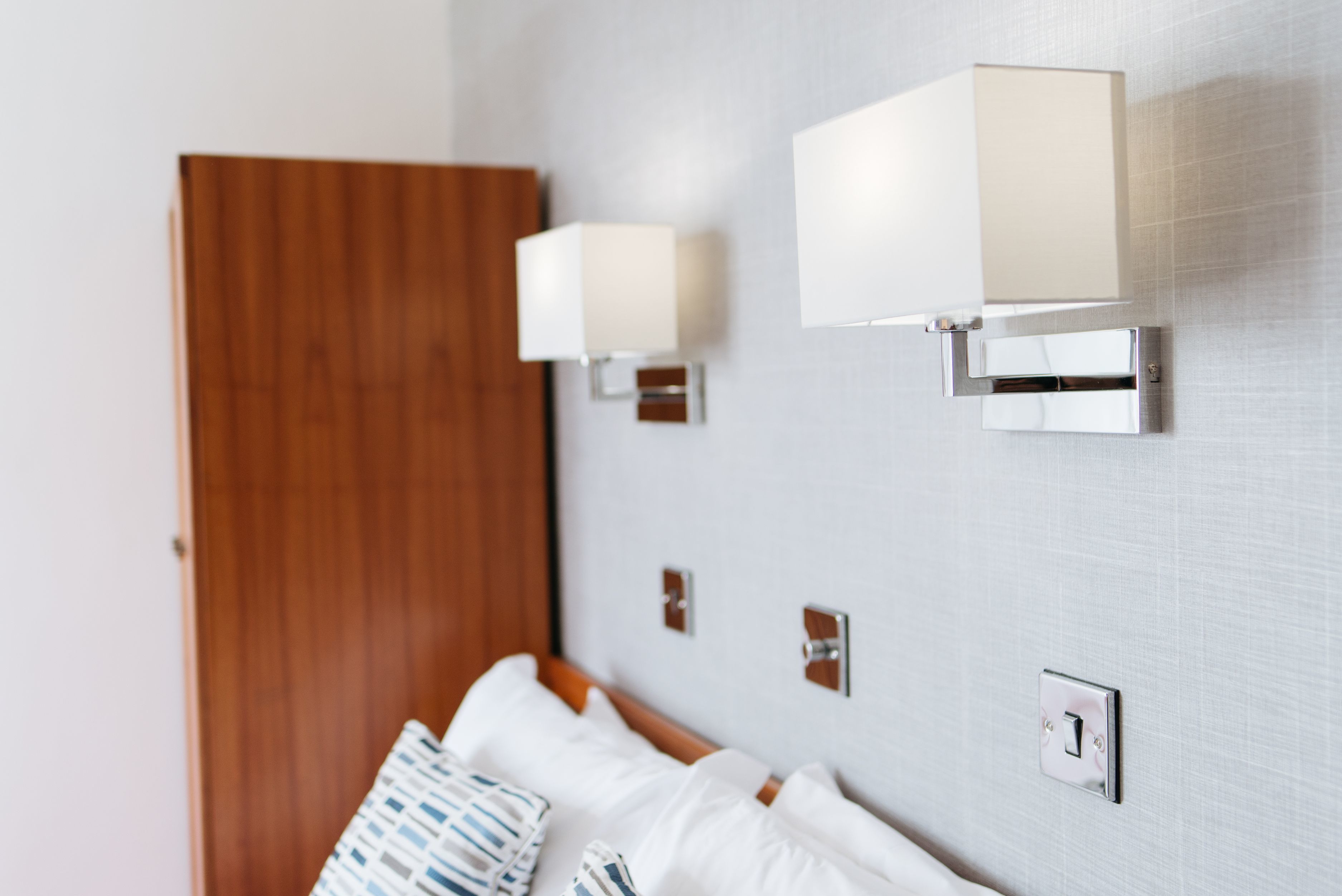 Quarto de hotel com porta de madeira, cama com roupa de cama branca e duas luminárias fixadas na parede.