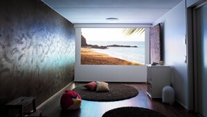 Quarto moderno com uma janela grande com vista para uma praia, um pequeno espaço de estar com almofadas e decoração minimalista.
