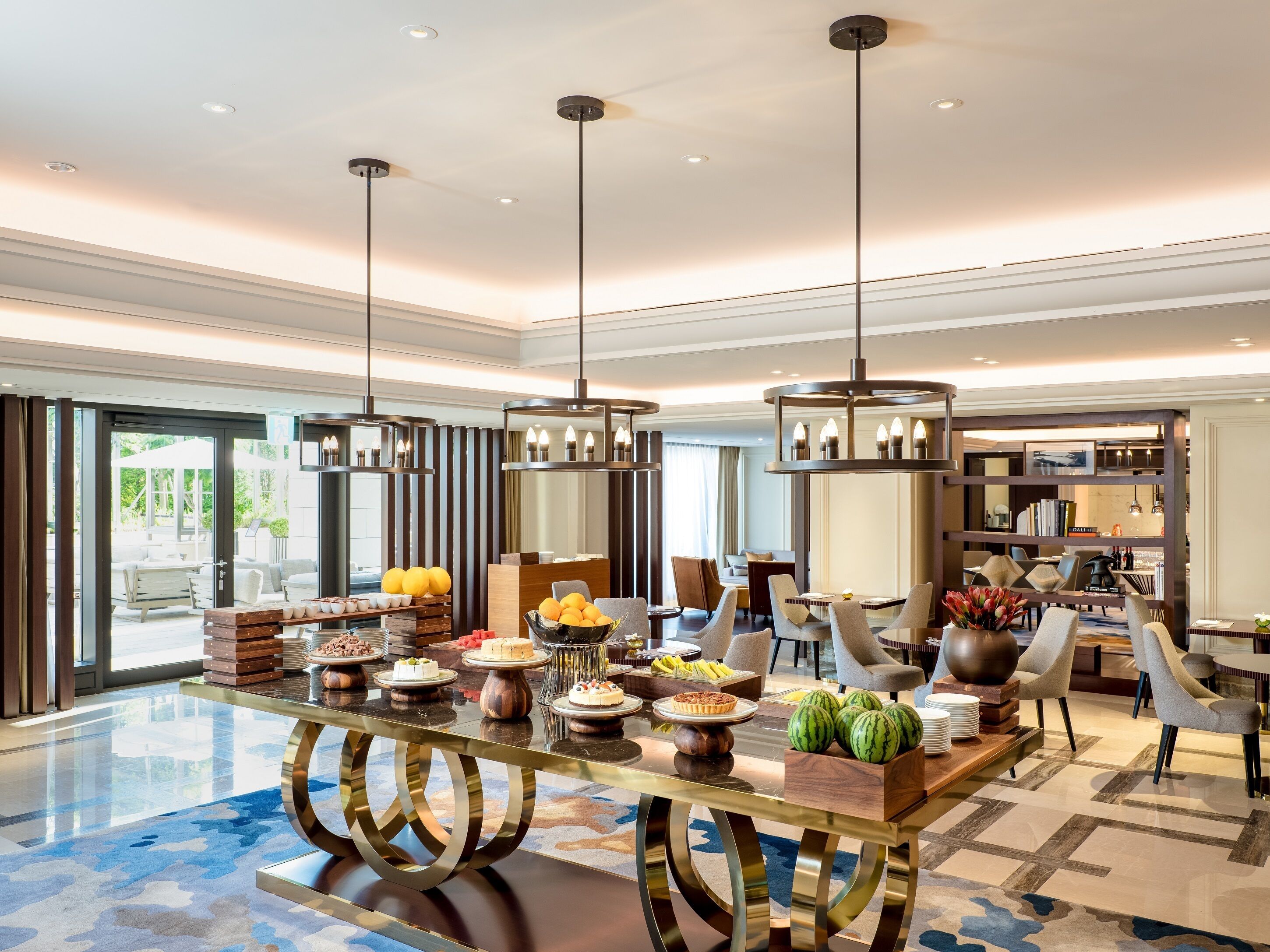 Um amplo saguão de hotel com uma mesa de jantar central, iluminação pendente moderna e vista para a área externa.