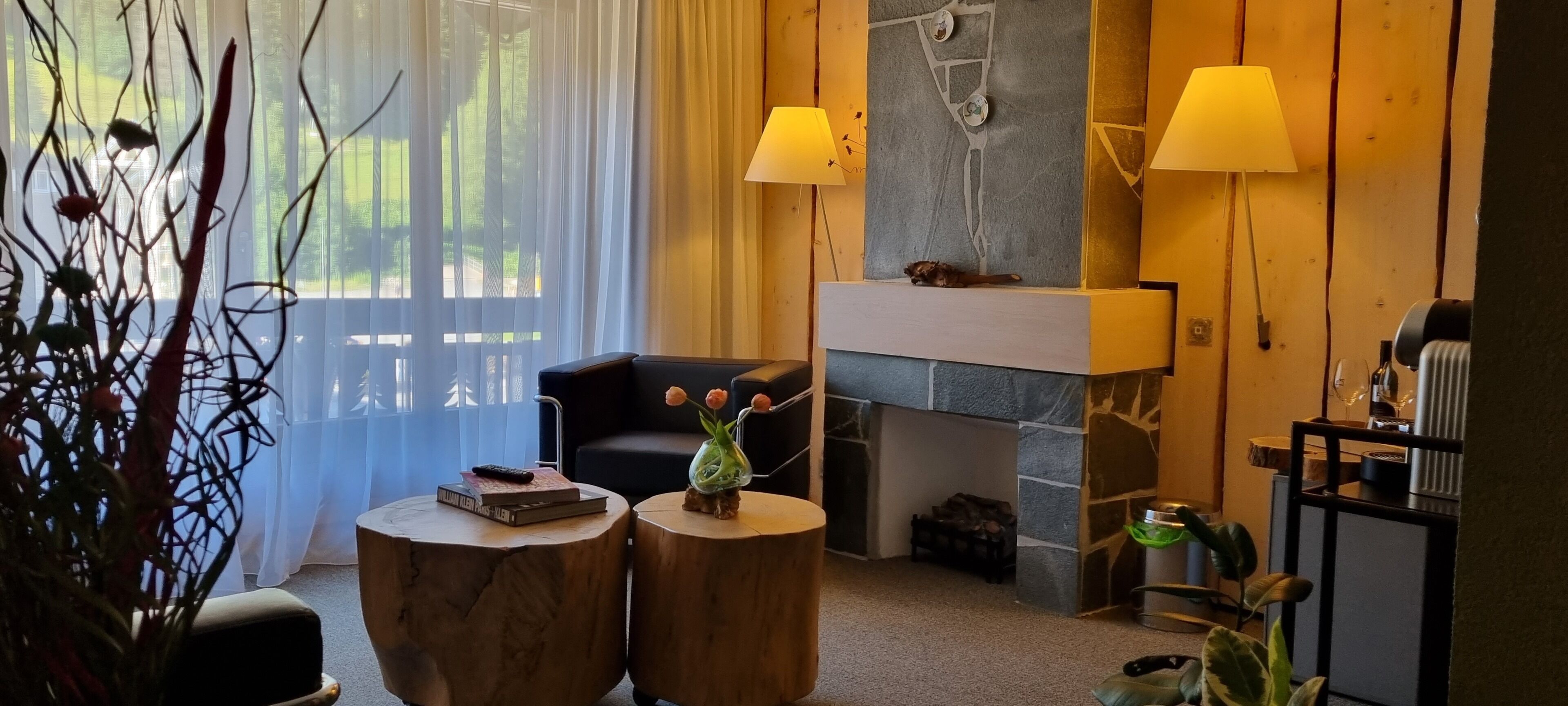 Uma sala de estar acolhedora com lareira, duas mesas redondas de madeira, um sofá confortável e vista para o exterior através de cortinas leves.
