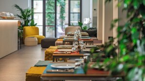 Uma sala de jantar moderna com mesas preparadas para refeição, um sofá amarelo e uma janela grande com vista para o verde.