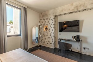 Quarto de hotel com cama, escrivaninha, cadeira, televisão e um padrão decorativo na parede.