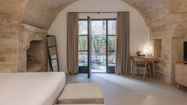 Quarto com arco de pedra, cama, escrivaninha com cadeira e vista para o exterior.