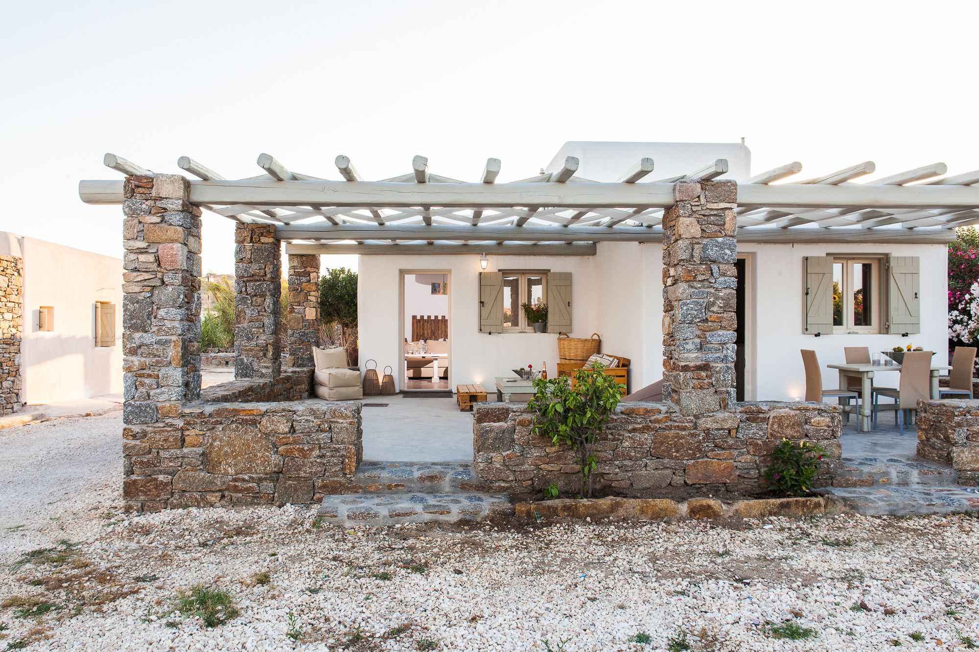 Um espaço ao ar livre com arco de pedra, uma pérgola, área de estar com mesa e cadeiras, e vista para o interior da casa.
