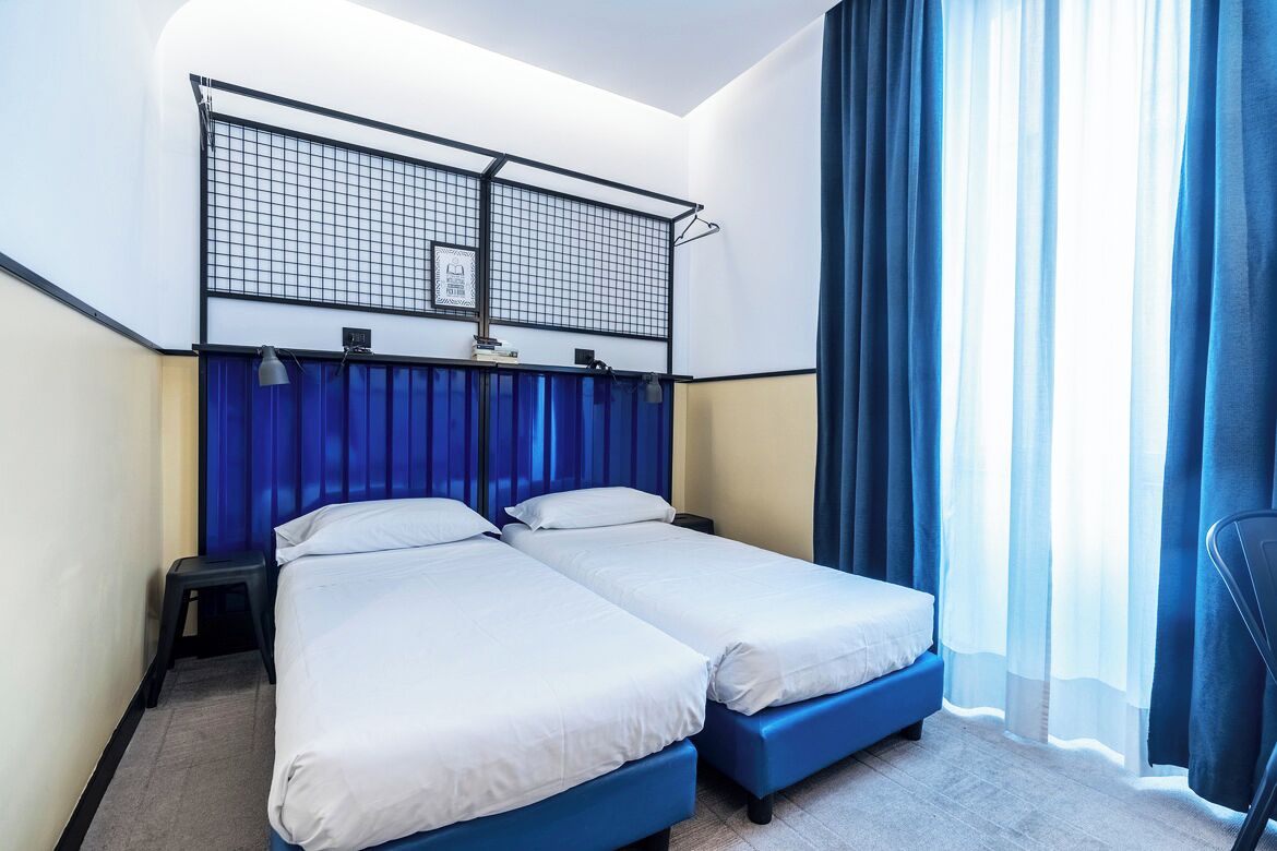 Quarto de hotel com duas camas de solteiro, uma cama beliche em cima, uma escrivaninha e uma cadeira.