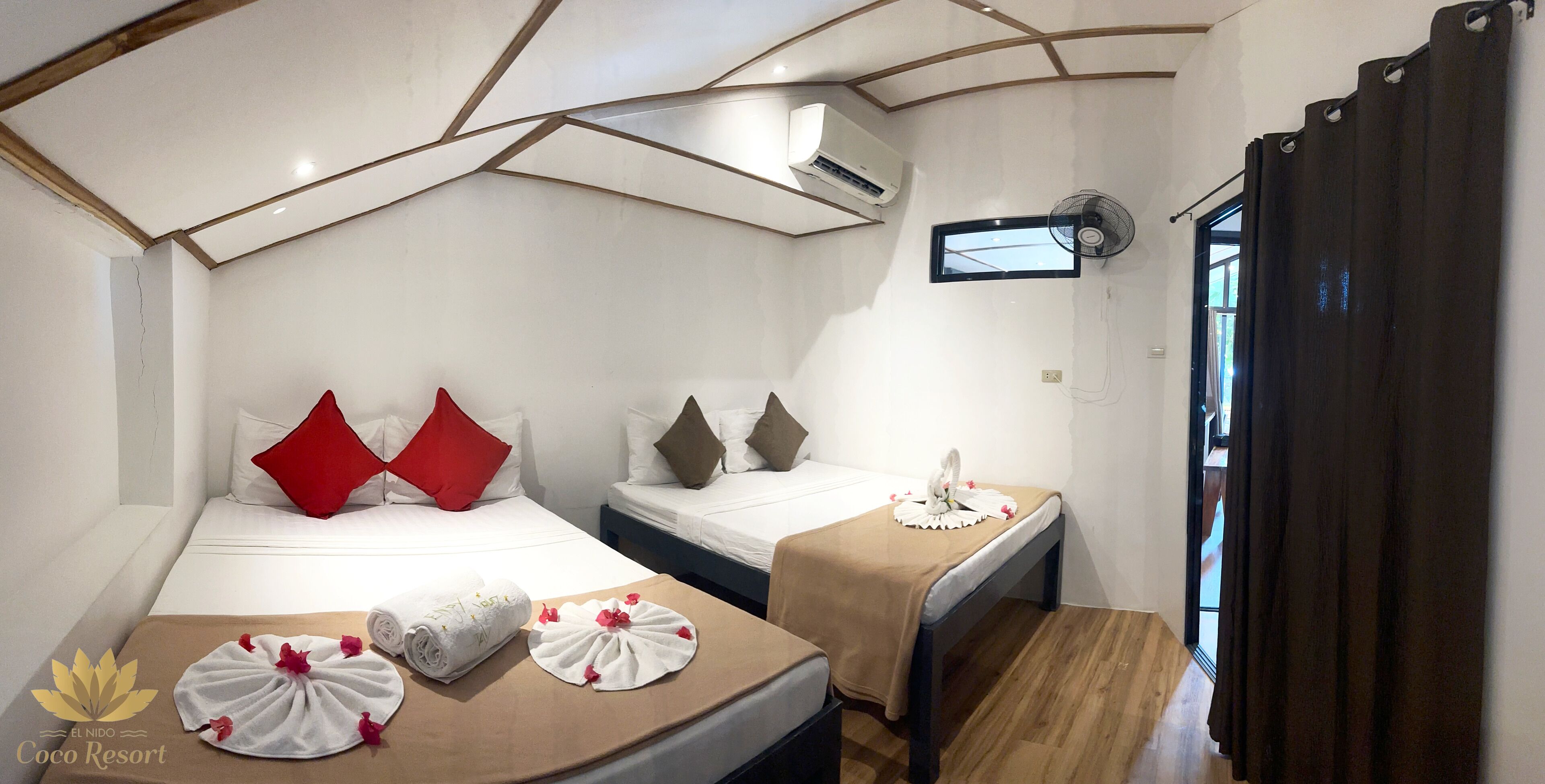Quarto de hotel com duas camas, uma janela, ar condicionado, um ventilador e uma mesa pequena com toalhas e flores.