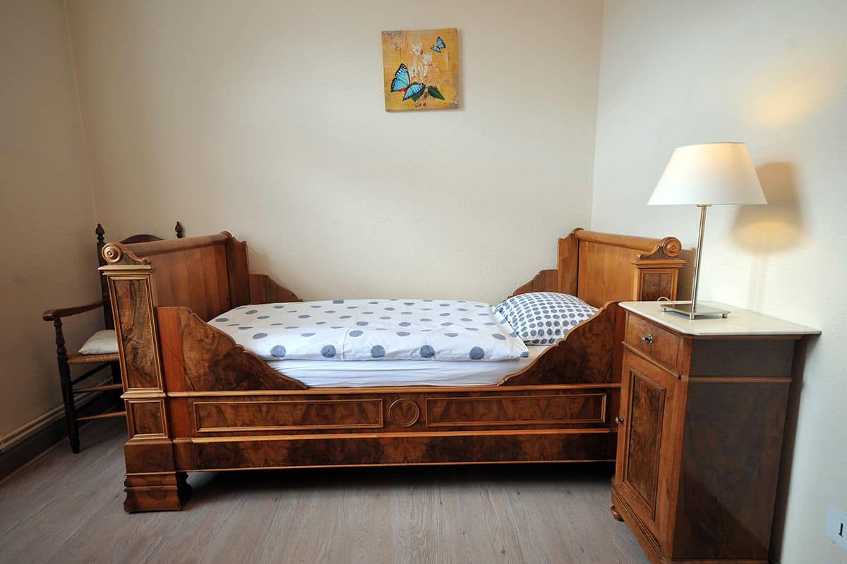 Uma cama de madeira com colchão branco e roupa de cama azul com bolinhas, um criado-mudo de madeira com um abajur e uma cadeira.