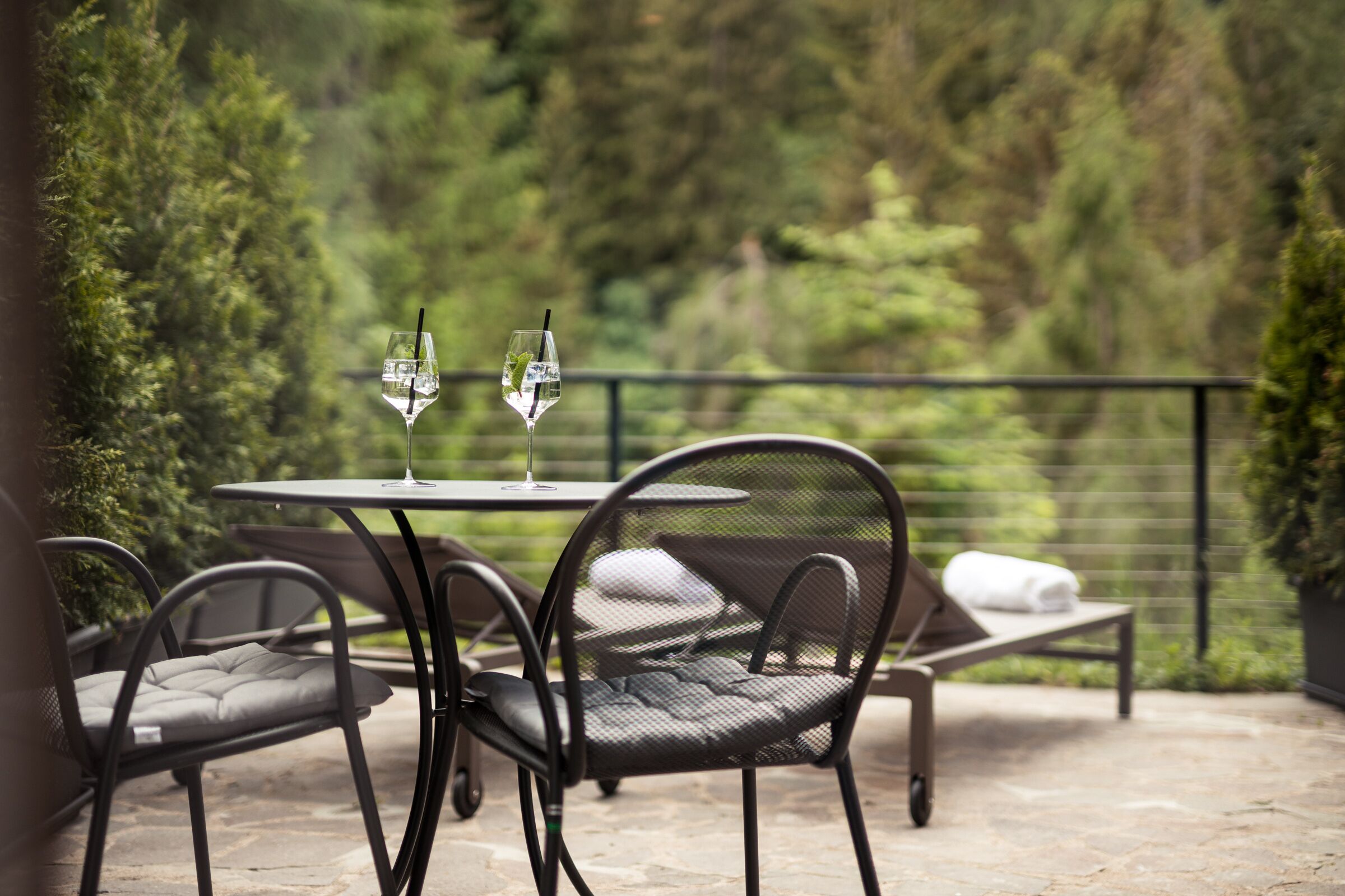 Uma área de pátio com mesa e cadeiras, uma poltrona de descanso e duas taças de vinho sobre a mesa.