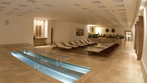 Um saguão de hotel com piscina, espreguiçadeiras, balcão de recepção e escadaria para os andares superiores.