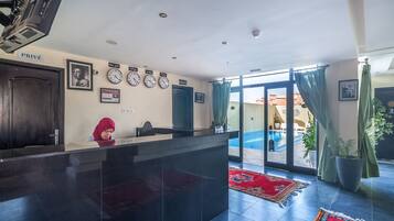 Uma área de recepção de hotel com um recepcionista, um balcão de atendimento, vista para a piscina e uma grande planta em vaso.