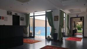 Um saguão de hotel com uma mesa de recepção, área de estar com vista para o mar e corredor que leva a outro quarto.
