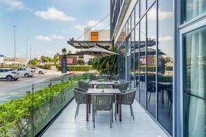 Uma área de refeições ao ar livre moderna com guarda-corpos de vidro, mesas e cadeiras, com vista para um estacionamento e um edifício com uma seção laranja marcante.