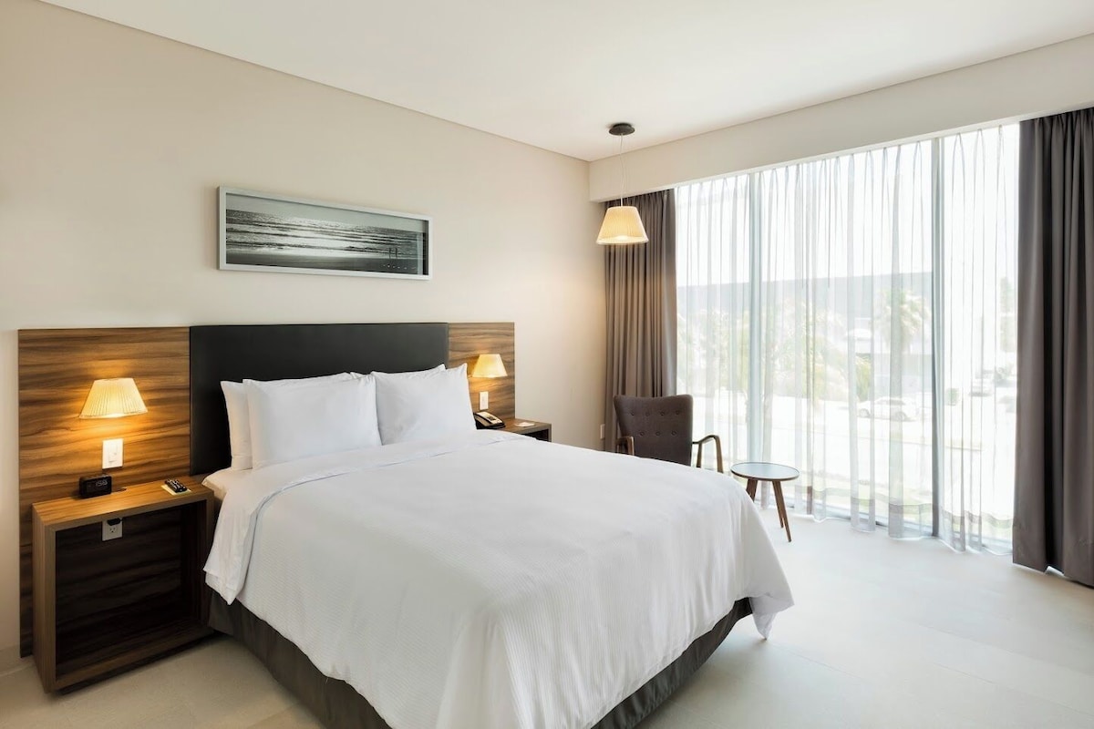 Quarto de hotel com uma cama, duas luminárias de cabeceira, uma cadeira, uma mesa pequena, um quadro na parede e uma janela grande com cortinas.