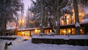 Área externa coberta de neve, com um edifício que possui grandes janelas e uma parede de pedra.
