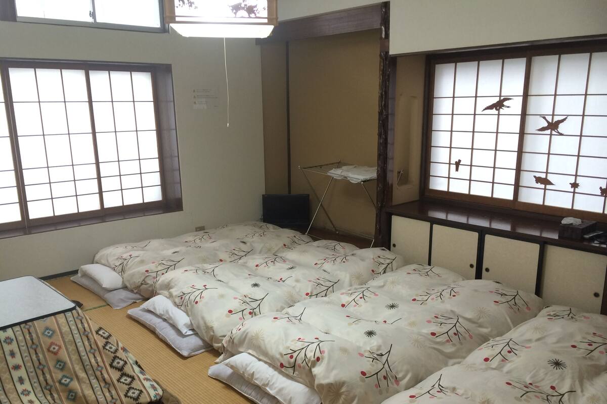 Quarto com duas camas, um futon, manta de futon, travesseiro de futon, colchão de futon, prateleira de madeira e janela com vista para pássaros.