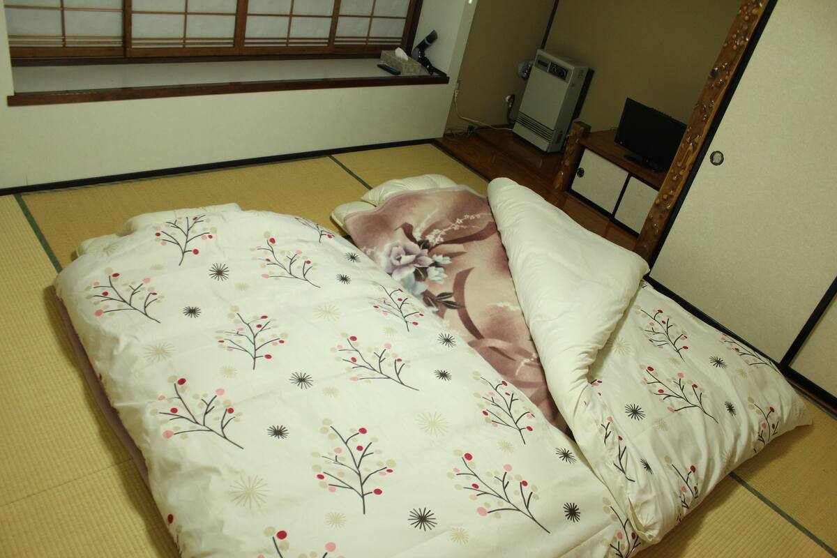 Um quarto japonês tradicional com tatames, futon com roupa de cama floral, armário de madeira com televisão e uma porta de correr.
