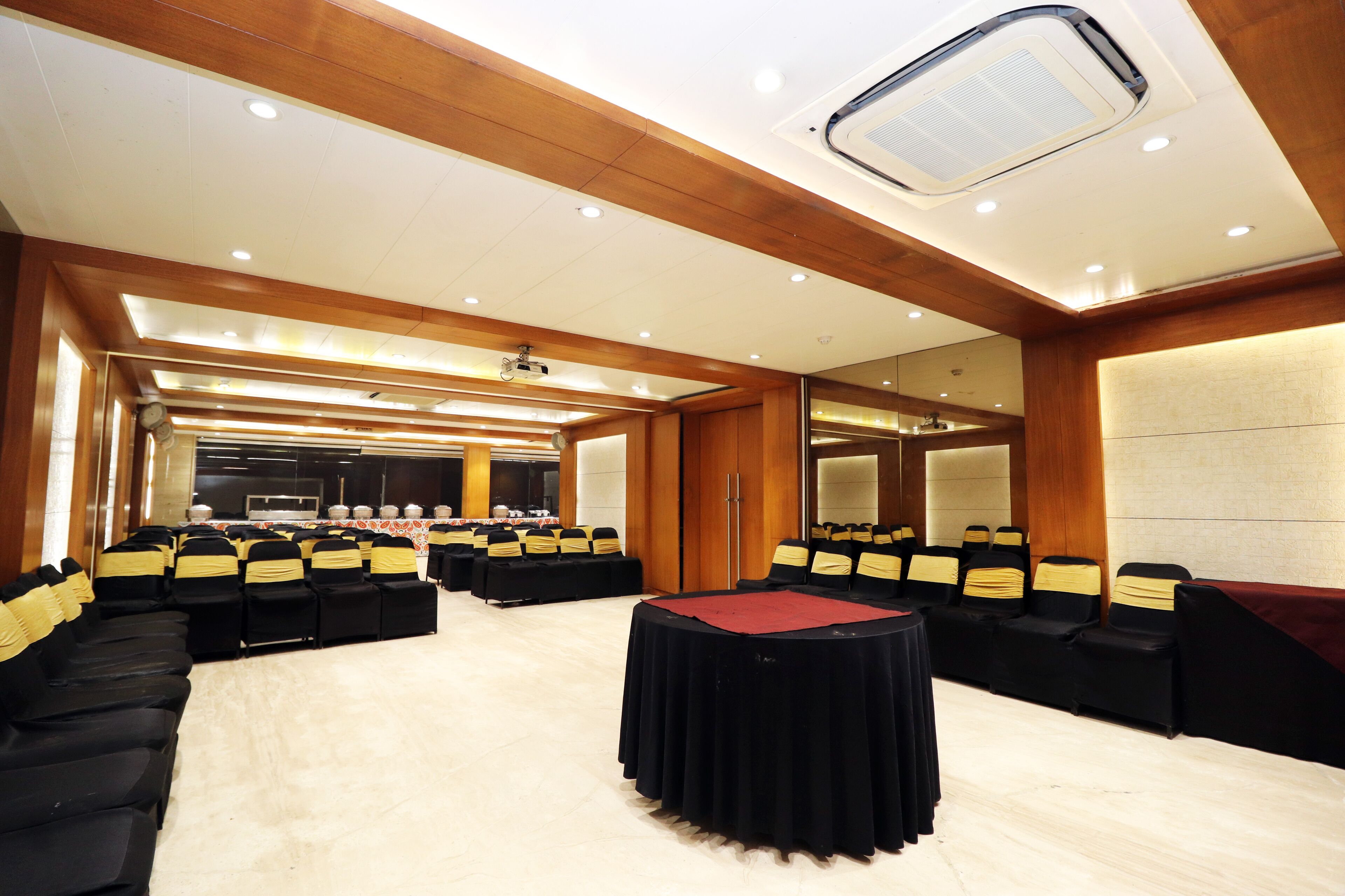 Uma sala de conferências com fileiras de cadeiras pretas com fitas amarelas, uma mesa redonda central com toalha de mesa vermelha e painéis de madeira no teto.