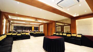 Uma sala de conferências com fileiras de cadeiras pretas com fitas amarelas, uma mesa redonda central com toalha de mesa vermelha e painéis de madeira no teto.