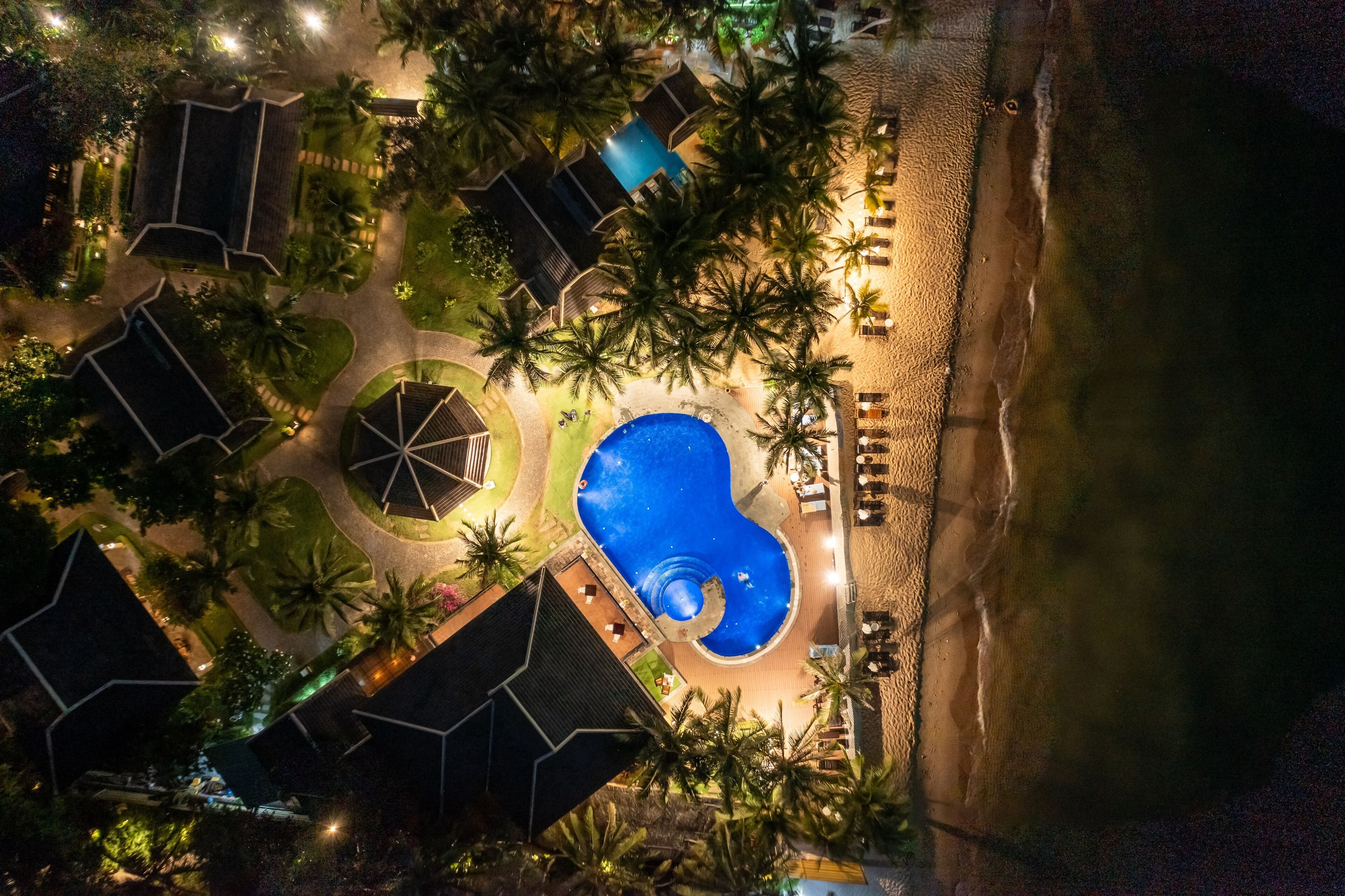 Vista aérea de um resort com piscina central, várias vilas para hóspedes e área de praia.