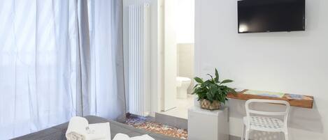 Quarto de hotel com cama, televisão na parede, secretária com cadeira e planta em vaso.