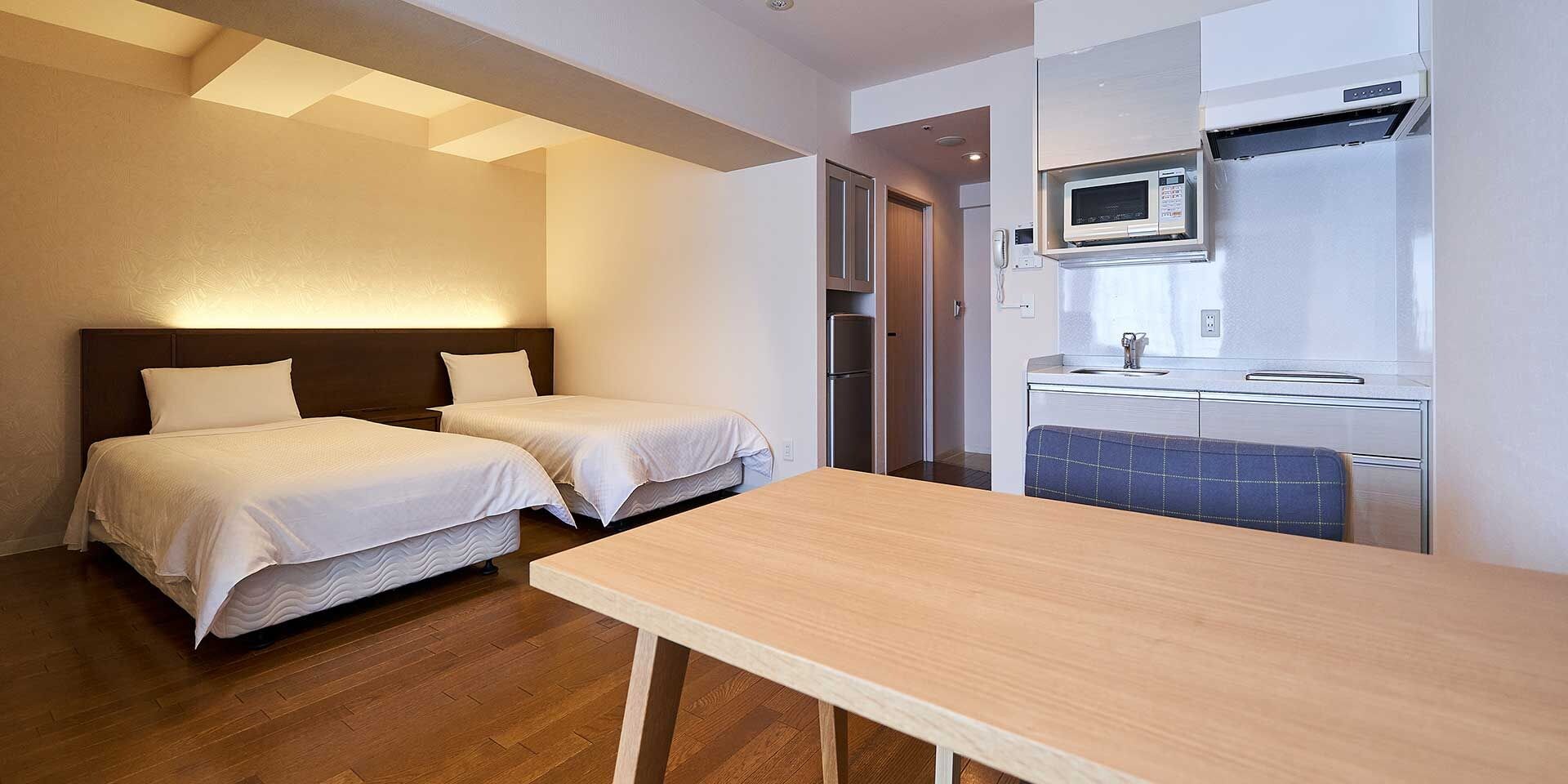 Quarto de hotel com duas camas, uma mesa de madeira, micro-ondas e uma pequena cozinha.