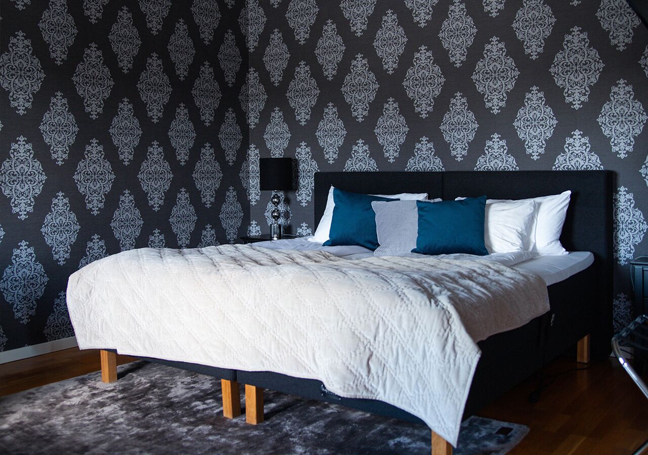 Quarto com cama, papel de parede estampado escuro, cabeceira preta e uma pequena mesa de cabeceira com abajur.