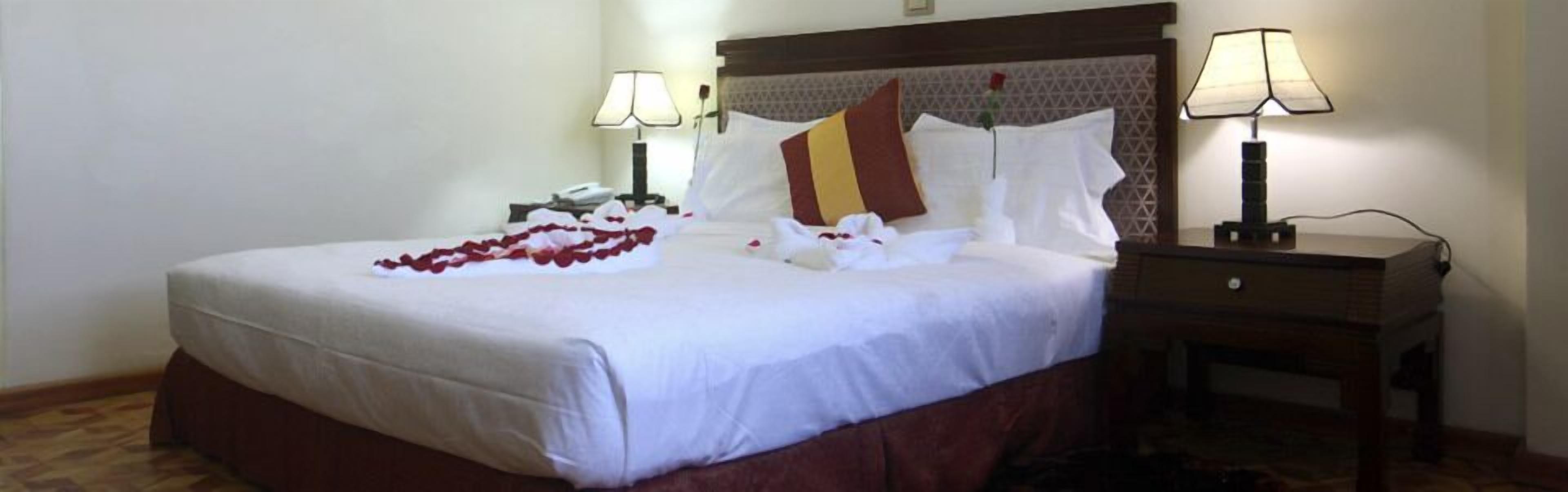 Cama bem arrumada com lençóis brancos, arranjo decorativo de rosas e duas luminárias de cabeceira.