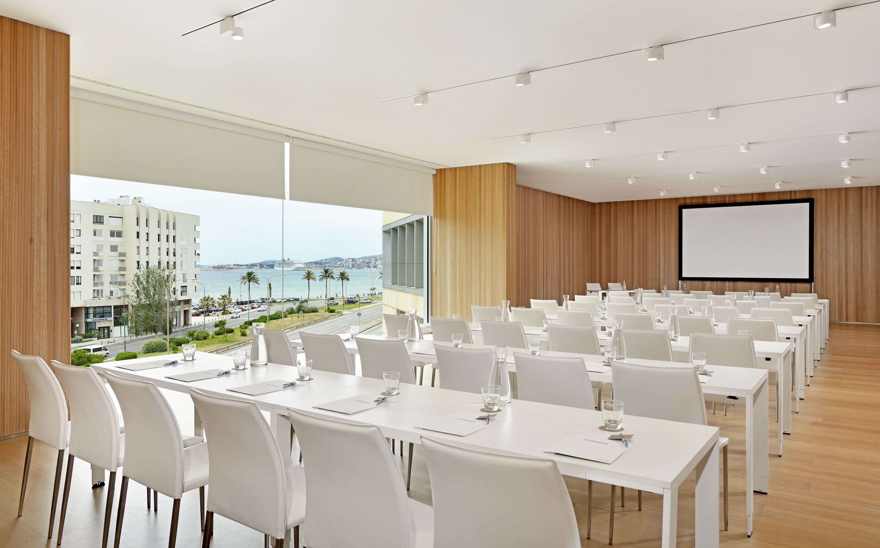 Uma sala de conferências com mesas e cadeiras brancas, uma tela grande e vista para o mar e edifícios.