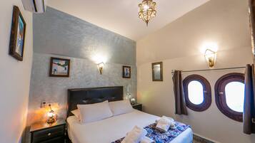 Quarto de hotel com cama, mesas de cabeceira, luminárias na parede, um candelabro e duas janelas com cortinas.