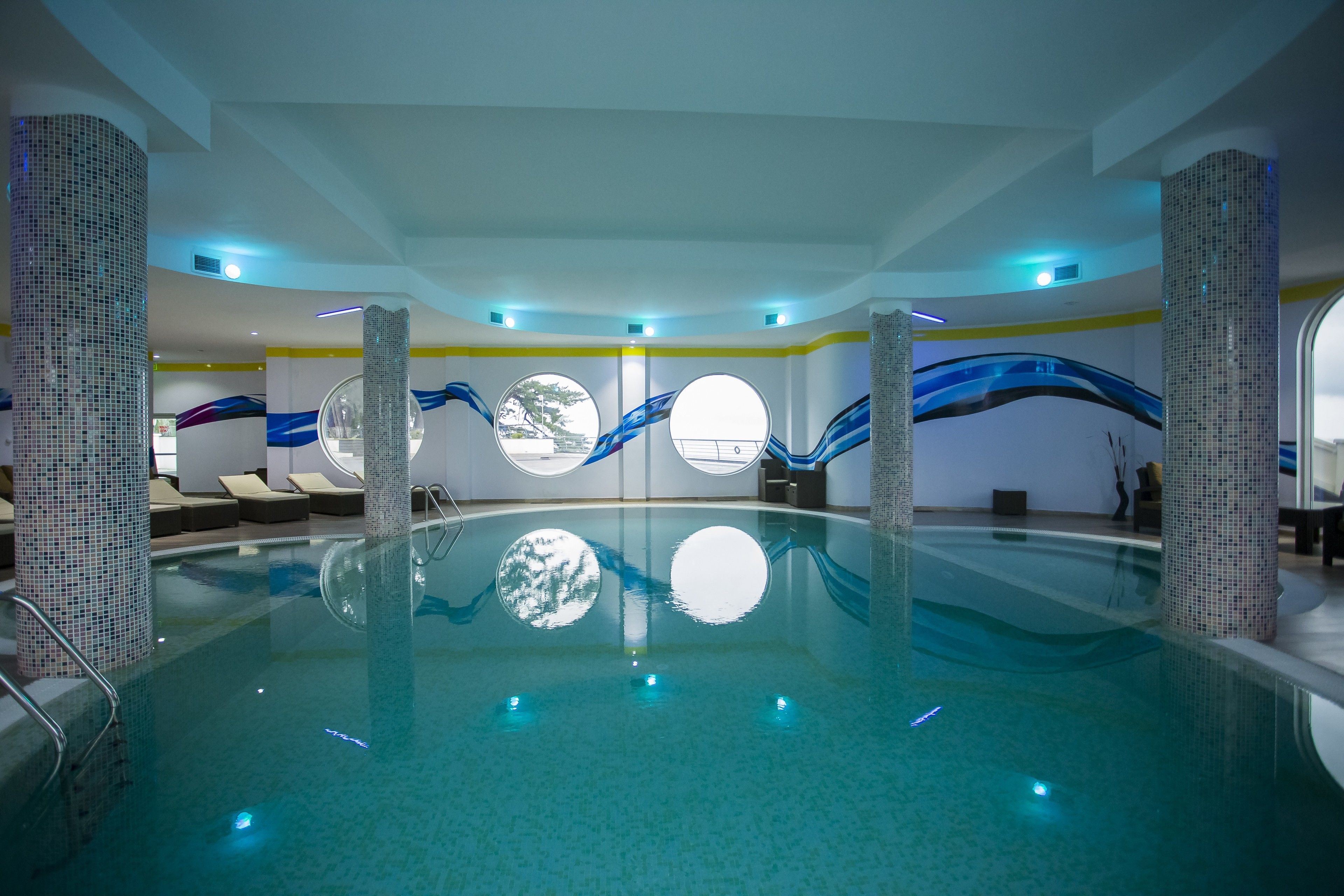 Uma piscina interior com água azul translúcida, rodeada por colunas com padrões em mosaico e um design moderno com arte em parede que lembra ondas azuis.