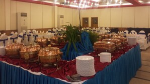 Uma mesa de banquete com travessas de cobre e pratos brancos, cercada por cadeiras brancas com fitas azuis.
