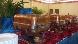 Salão de festas com várias estações de buffet, uma grande planta em vaso e teto com iluminação embutida.