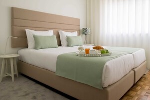 Uma cama bem arrumada com um cobertor verde, um tabuleiro com frutas e um copo de sumo de laranja, e um abajur.