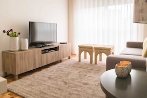 Uma sala de estar moderna com uma televisão de tela plana em um armário de madeira, um tapete bege, um banquinho estampado e uma pequena mesa com xícaras decorativas.