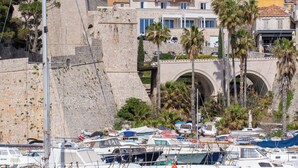 Uma vila costeira com edifícios históricos de pedra, um porto com barcos e uma imponente muralha de pedra.