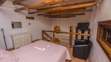 Um quarto com cama, cômoda, poltrona, televisão e vigas de madeira no teto.