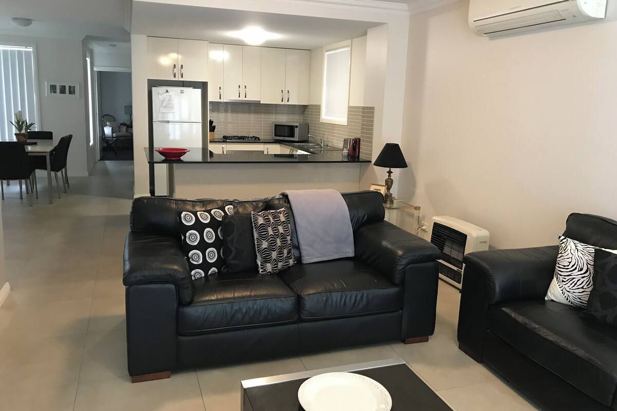 Uma sala moderna com um sofá de couro preto, uma área de jantar com mesa e cadeiras brancas e uma cozinha com armários brancos e micro-ondas.