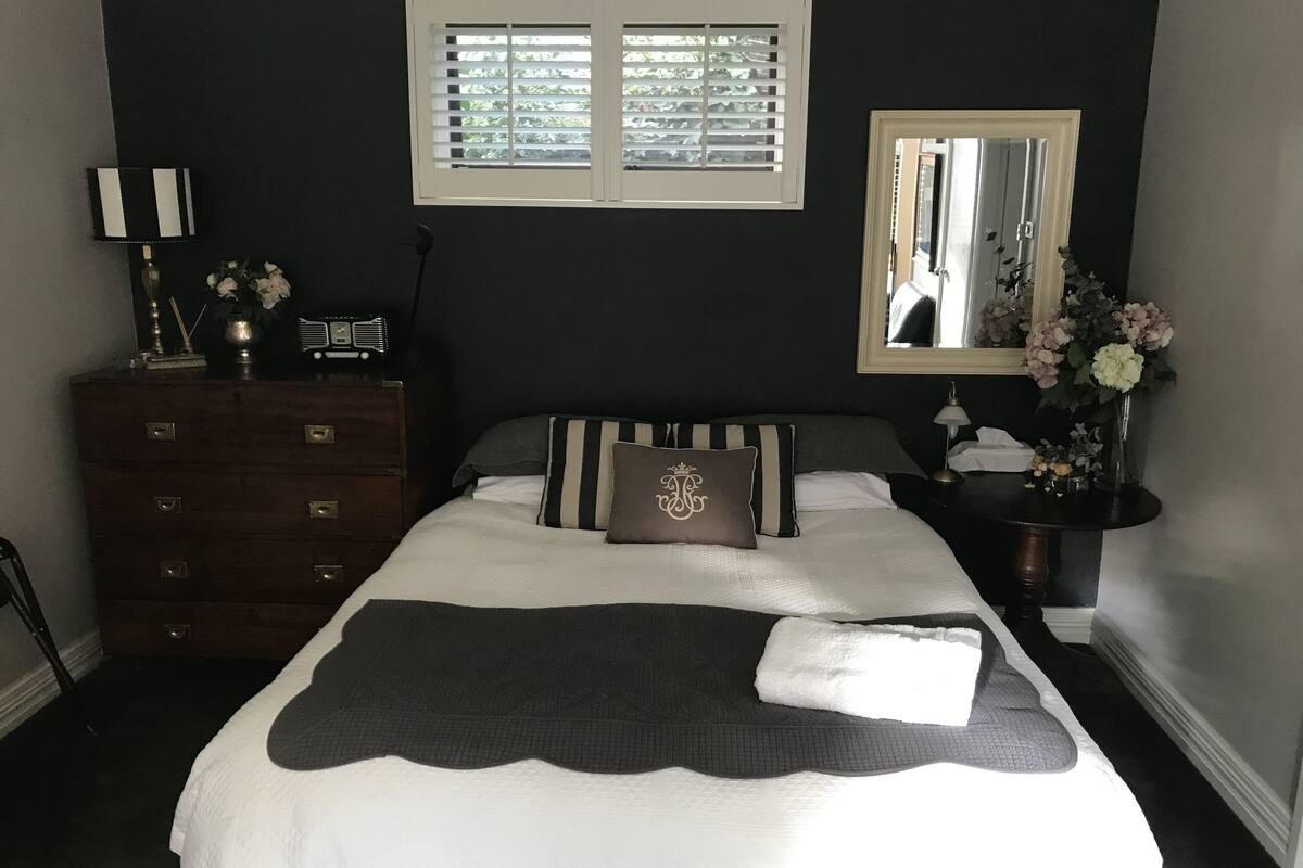 Quarto com cama, penteadeira, janela, espelho e mesa de cabeceira com flores.