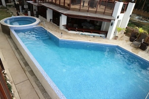 Uma piscina retangular com borda em azulejo, adjacente a um edifício com área de estar coberta ao ar livre.