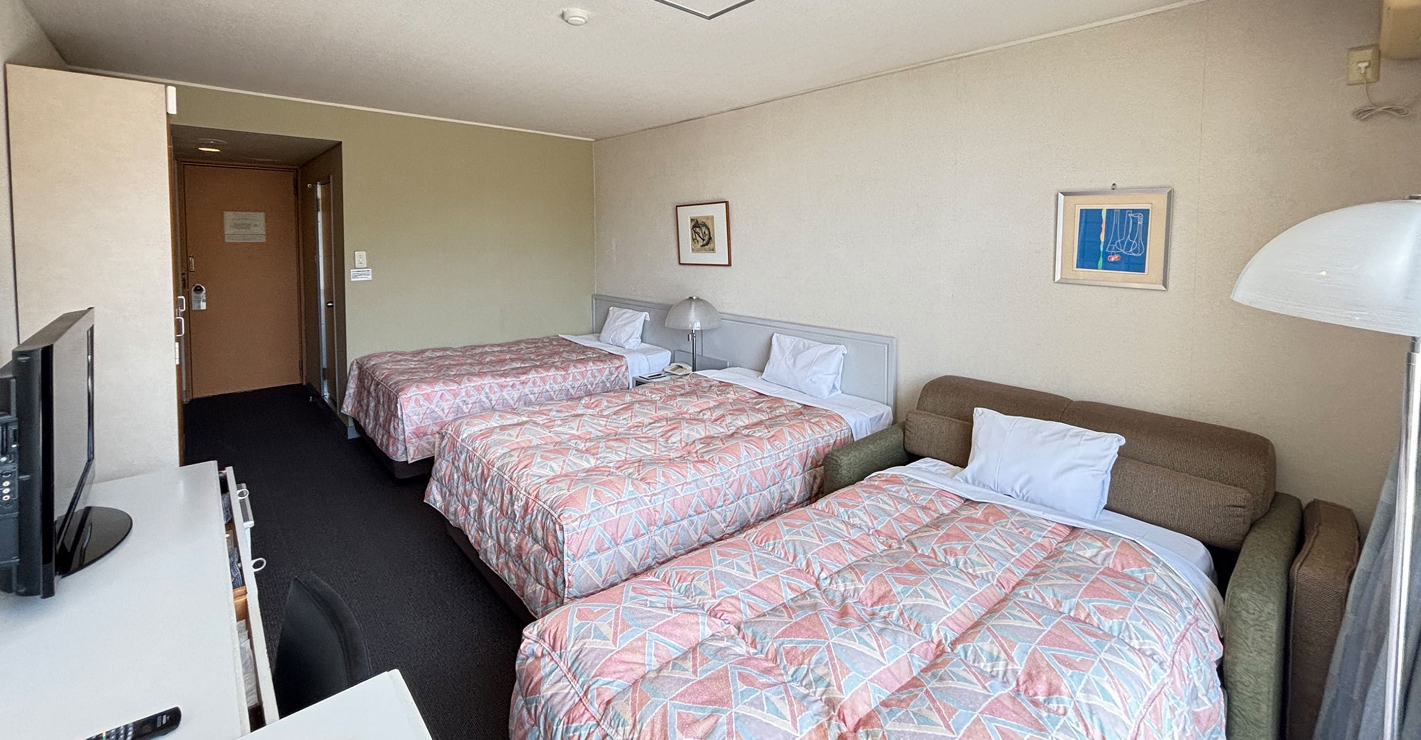 Quarto de hotel com duas camas, uma escrivaninha, uma televisão e uma porta que dá para outro cômodo.