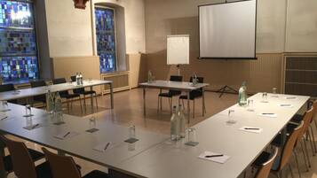 Uma sala de conferências com mesas compridas, cadeiras, tela de projetor e janelas de vitrais.