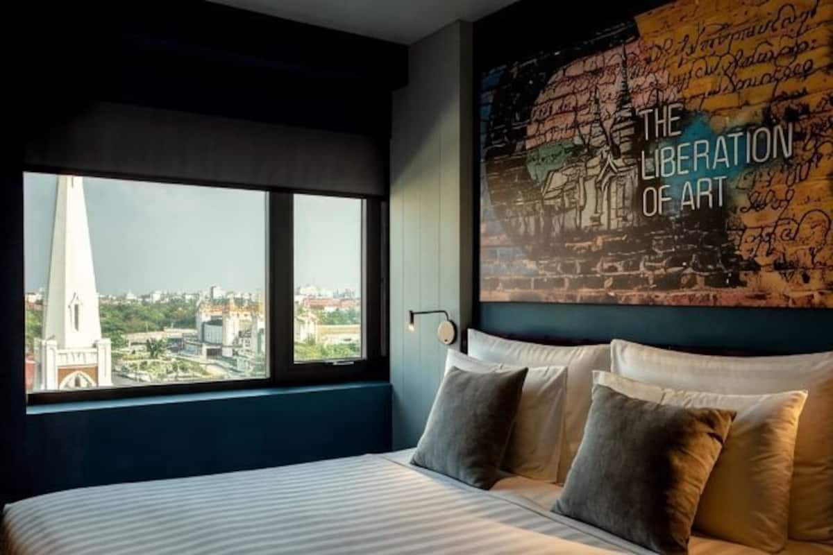 Quarto de hotel com uma cama grande, vista para a cidade através de uma janela e um mural na parede.