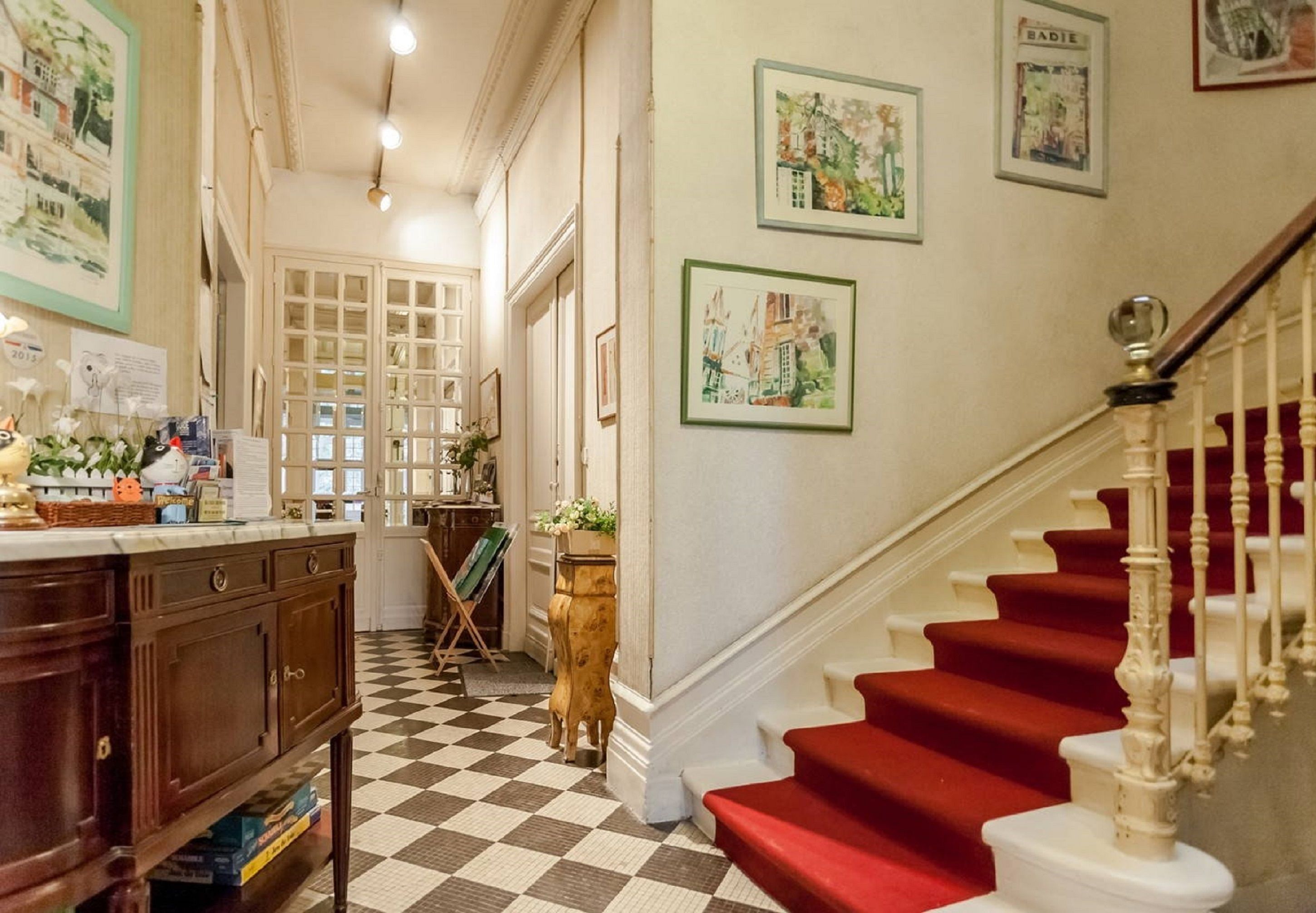 Um corredor com piso xadrez, escada com carpete vermelho e obras de arte emolduradas nas paredes.