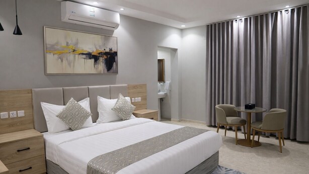 Quarto de hotel moderno com uma cama grande, mesas de cabeceira, uma escrivaninha com cadeiras, banheiro e uma janela ampla com cortinas.
