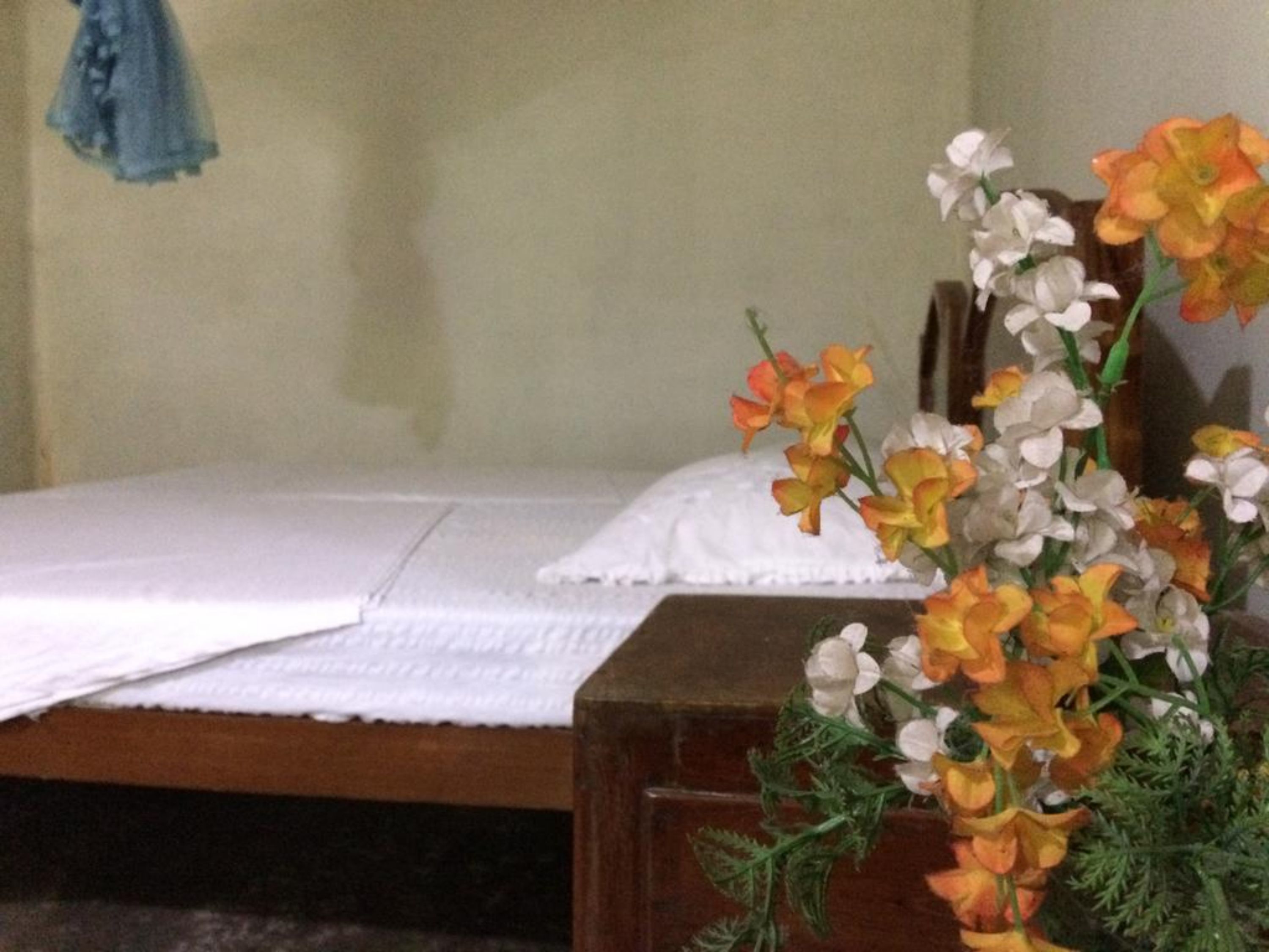Uma cama com roupa de cama branca e cabeceira de madeira, uma pequena mesa com um vaso de flores, e uma toalha azul pendurada na parede.