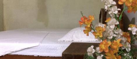 Uma cama com roupa de cama branca e cabeceira de madeira, uma pequena mesa com um vaso de flores, e uma toalha azul pendurada na parede.