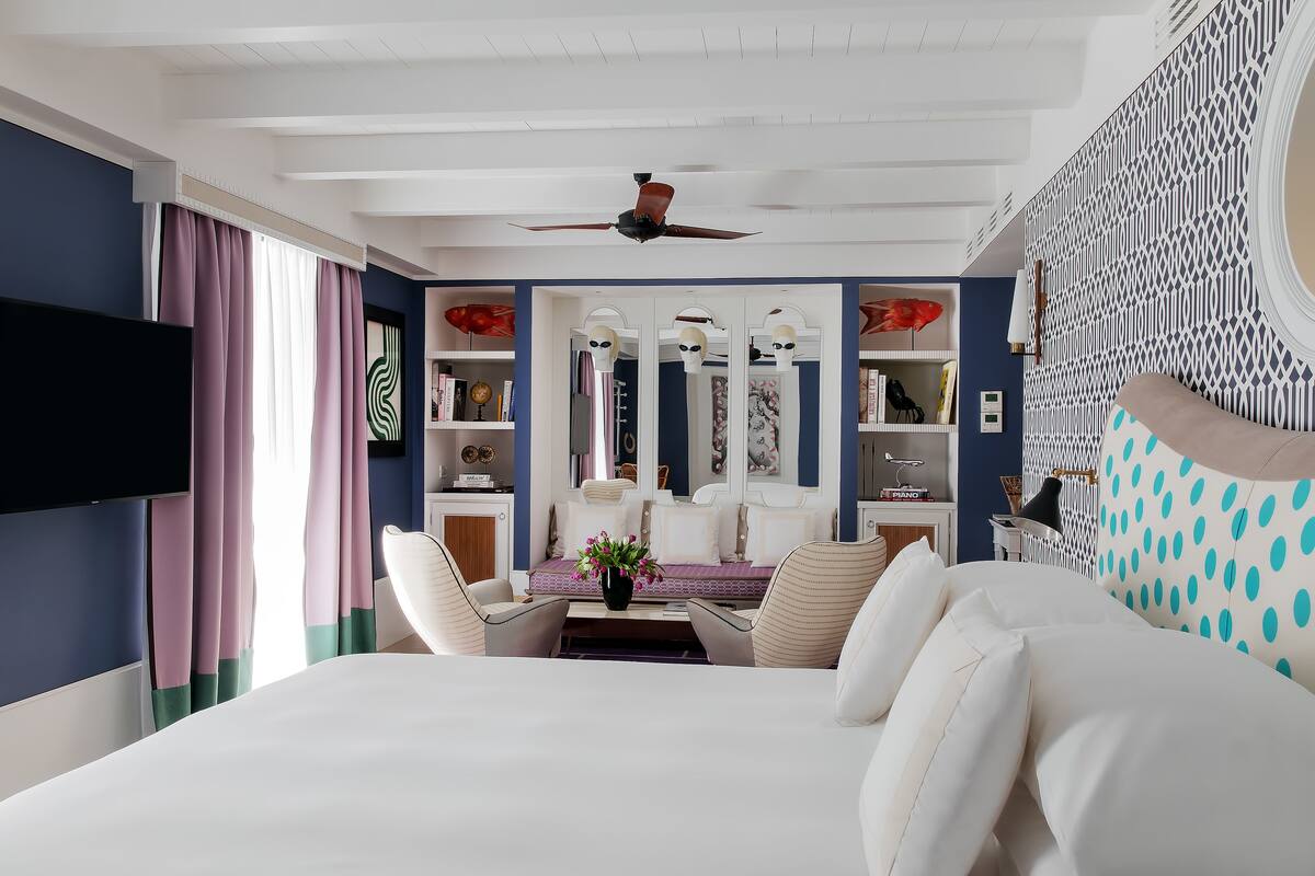 Quarto de hotel moderno com uma cama grande, uma escrivaninha com cadeira, um ventilador de teto e uma TV na parede.